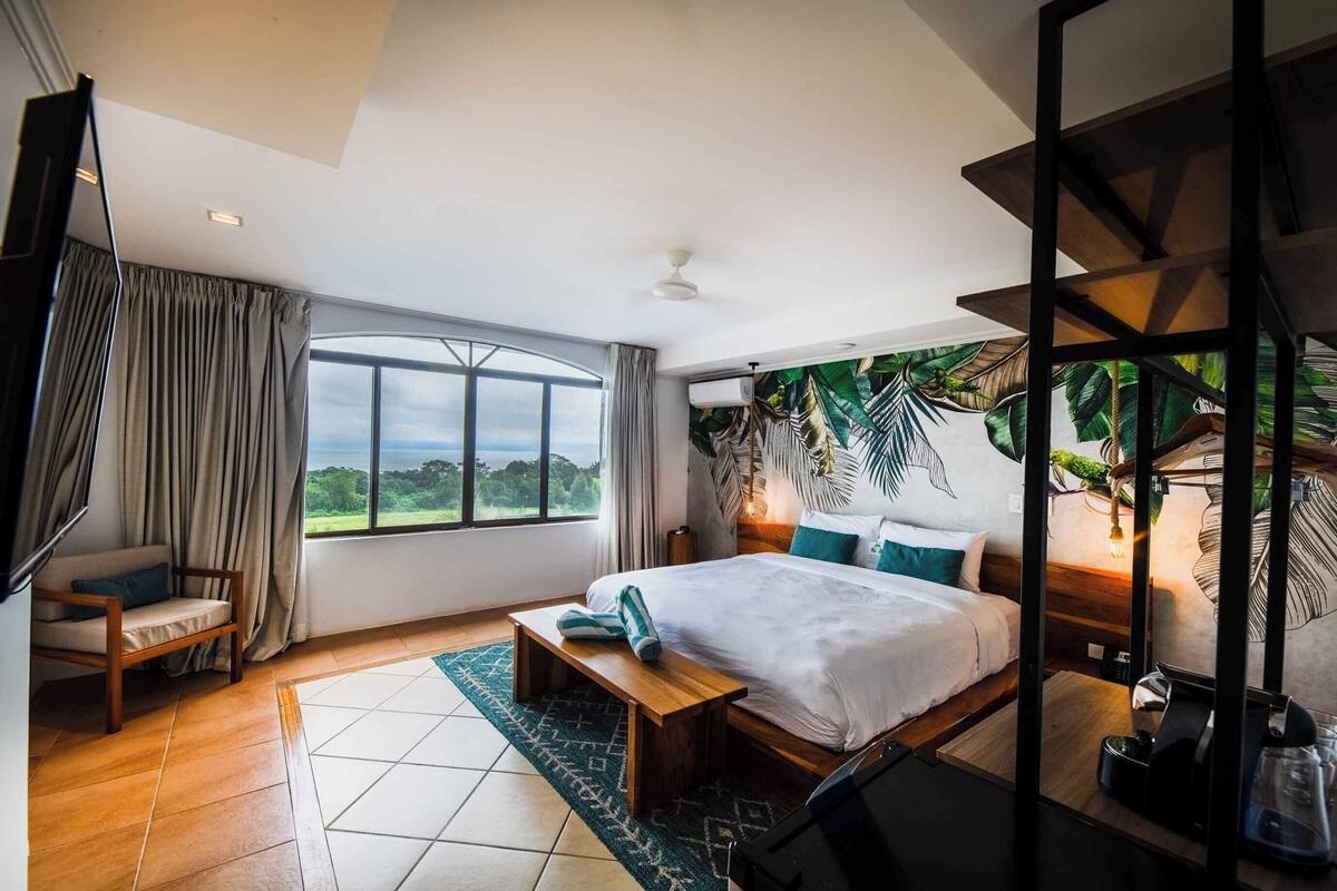 Quarto com cama grande, um banquinho, um sofá e vista para o exterior.