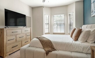 Quarto com uma cama, televisão sobre um aparador de madeira e janelas com persianas.
