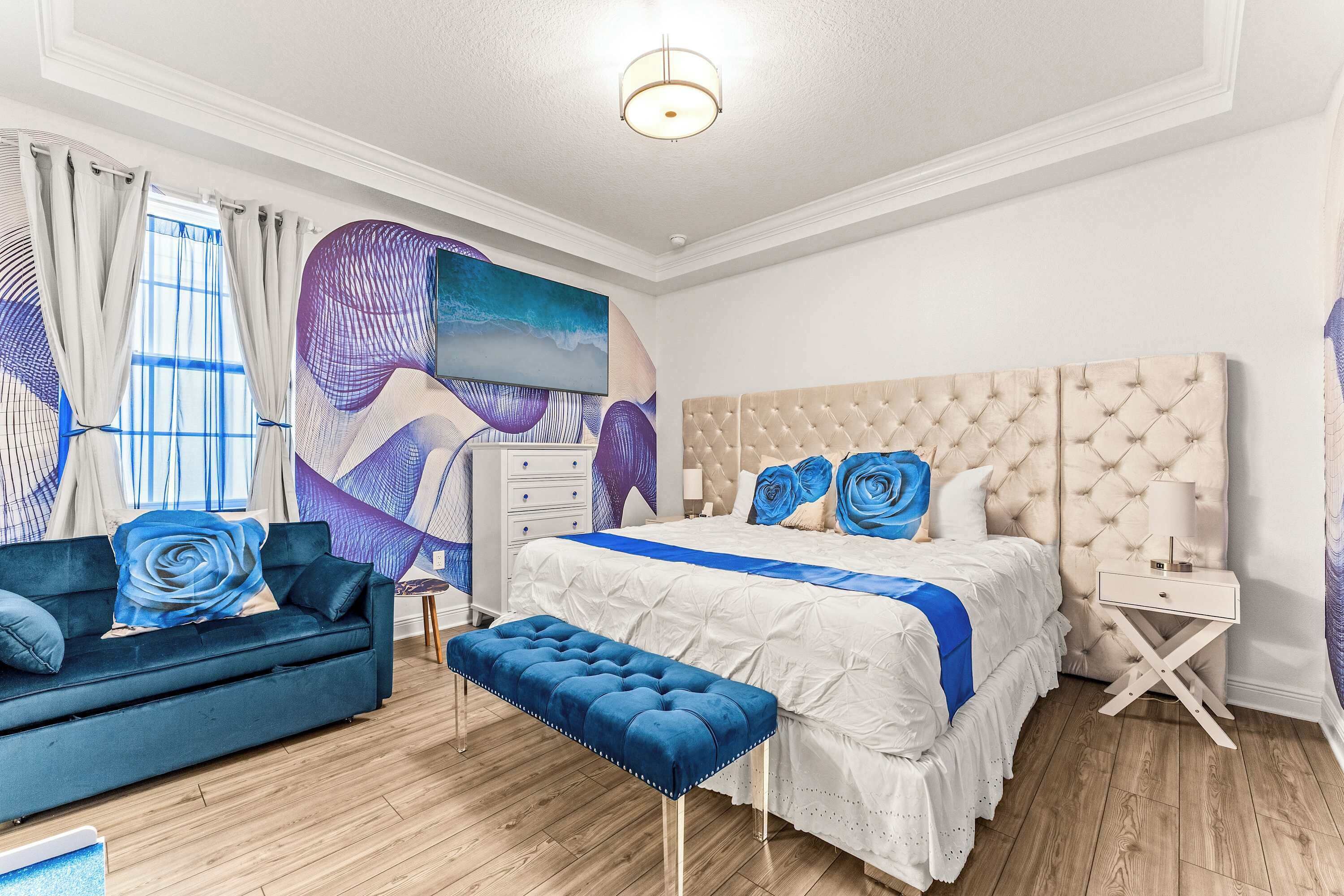 Quarto moderno com cama de cabeceira estofada, um banquinho azul, um sofá azul e um quadro abstrato grande na parede.