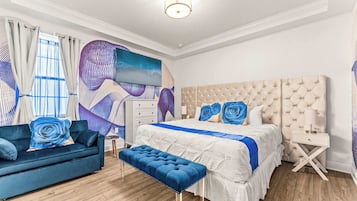 Quarto moderno com cama de cabeceira estofada, um banquinho azul, um sofá azul e um quadro abstrato grande na parede.