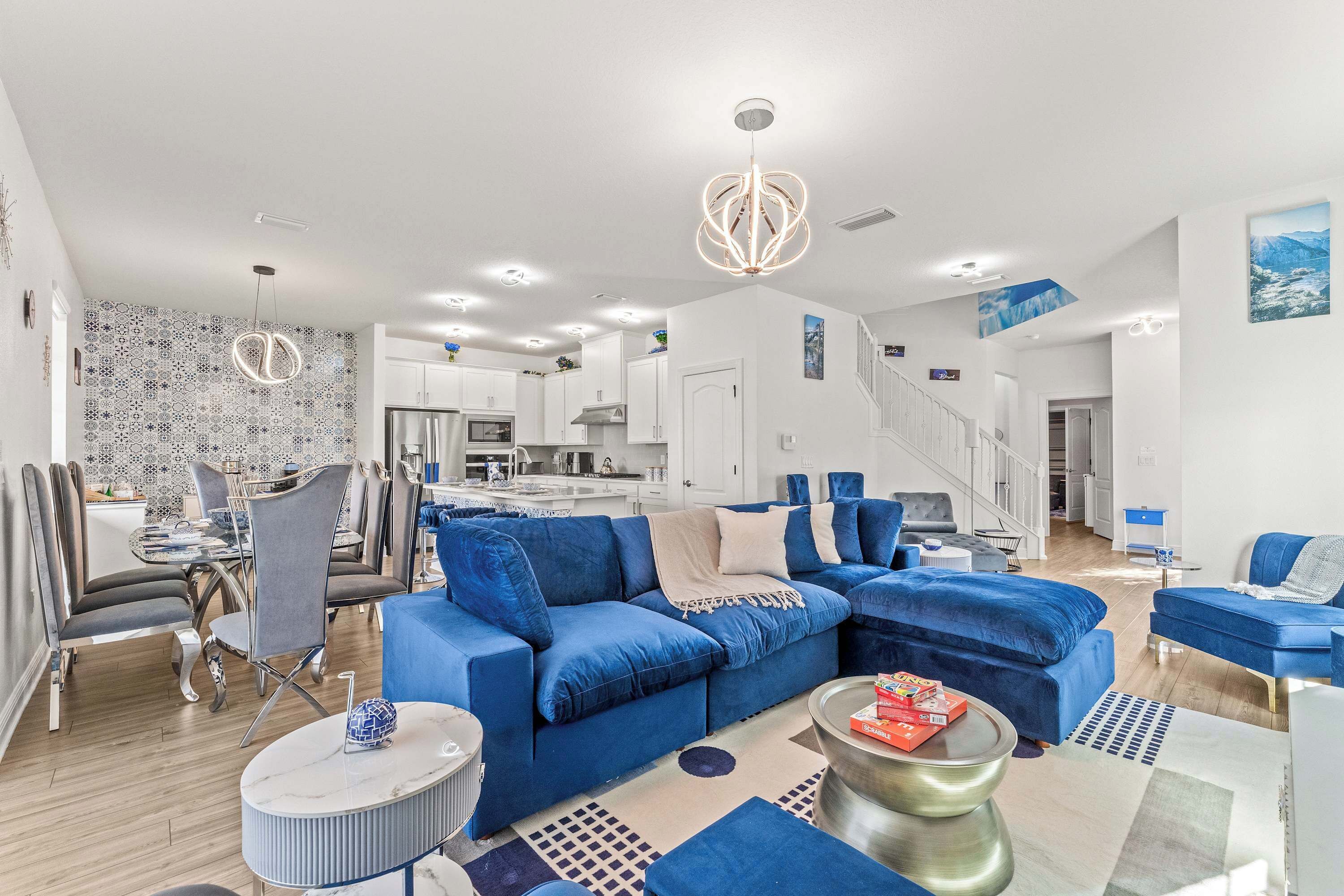 Uma sala de estar moderna com um sofá seccional azul, uma área de jantar com mesa de vidro e uma cozinha ao fundo.