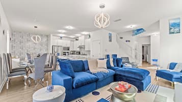 Uma sala de estar moderna com um sofá seccional azul, uma área de jantar com mesa de vidro e uma cozinha ao fundo.