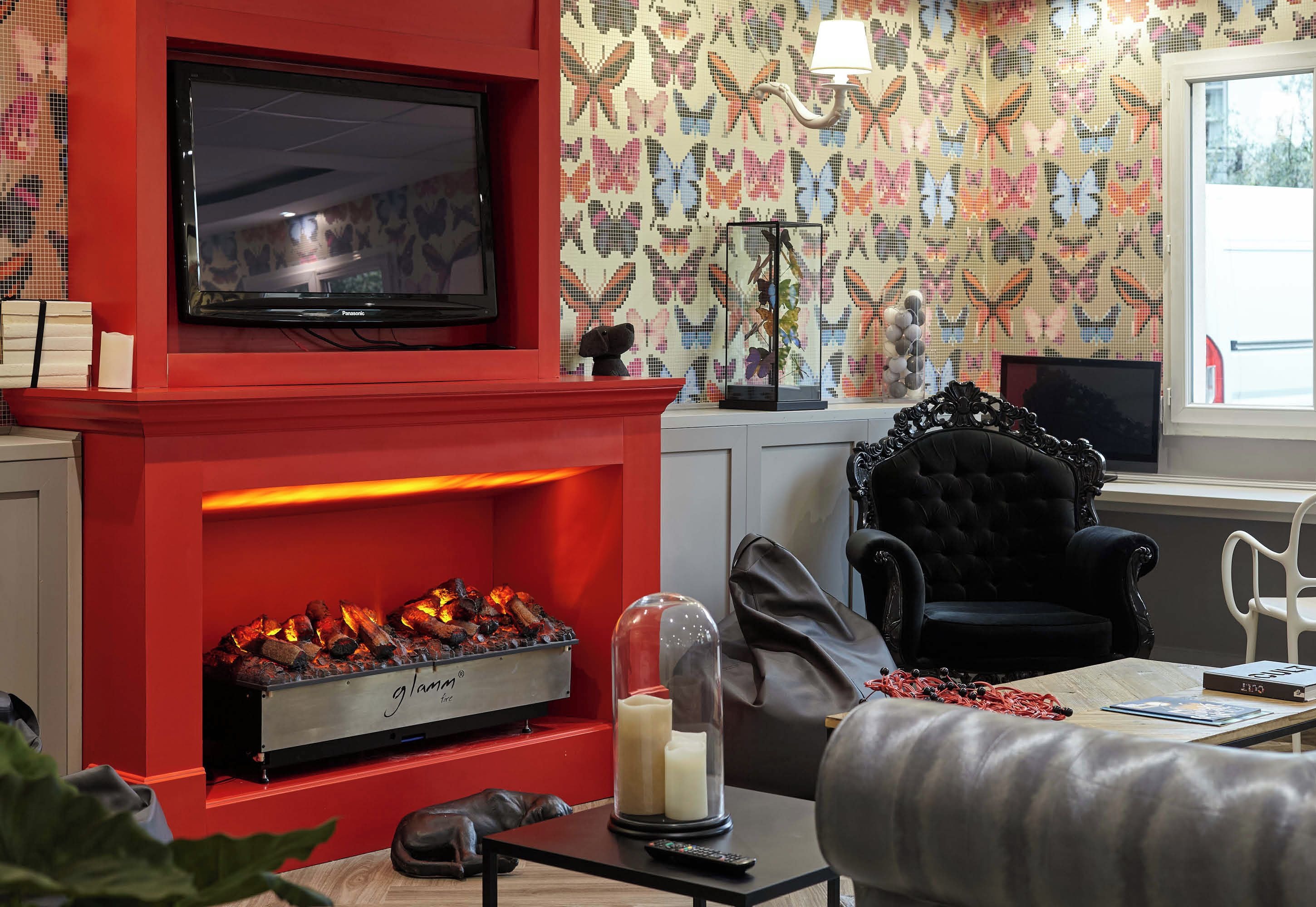 Uma sala de estar com lareira vermelha, televisão de tela plana, papel de parede estampado, poltrona preta e cadeira branca.