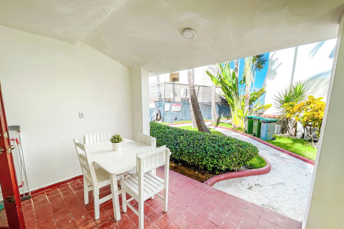 Quarto com uma mesa e cadeiras brancas, uma pequena planta em vaso, e vista de um jardim bem cuidado com um caminho, árvores e um edifício ao fundo.