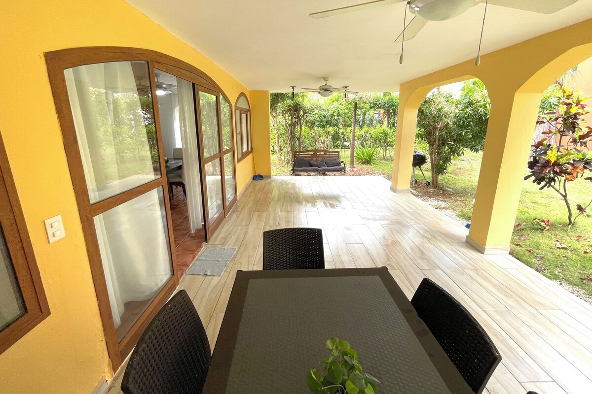 Uma varanda coberta com mesa e cadeiras, janelas amplas e um ventilador de teto.