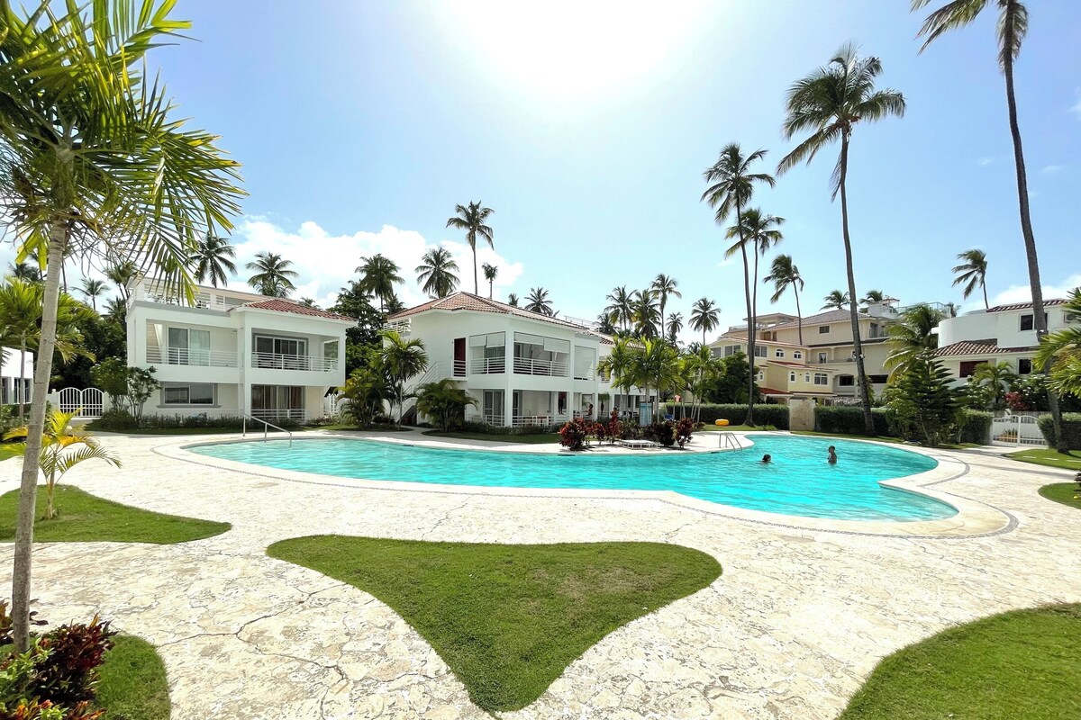 Piscina com área gramada em formato de coração, cercada por palmeiras e edifícios modernos.