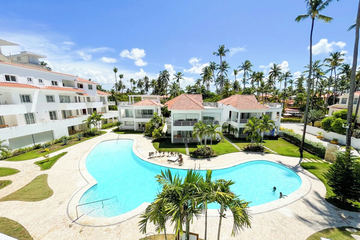 Um resort com uma grande piscina, vários edifícios brancos com telhados de terracota e palmeiras.