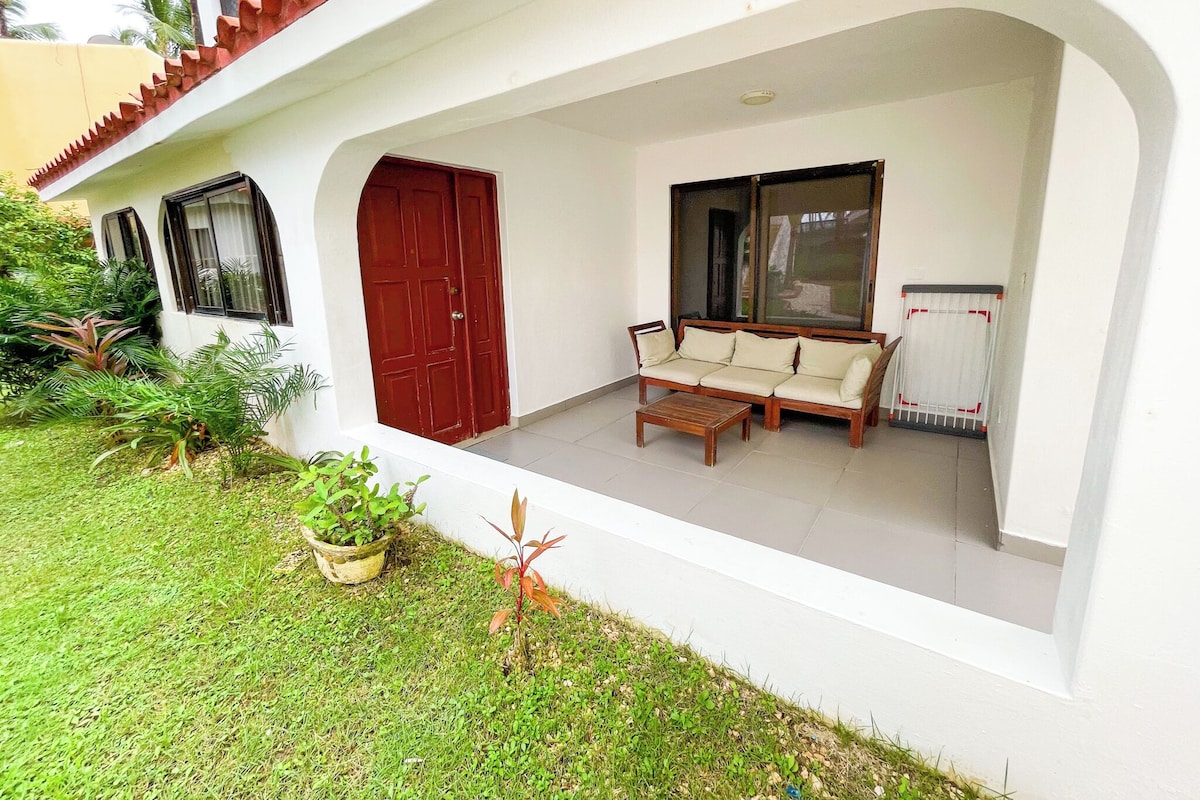 Uma área de estar coberta ao ar livre, com um sofá e uma mesa, uma porta vermelha e uma janela com vista para o exterior.