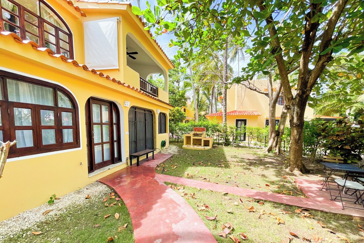 Um edifício amarelo com um caminho vermelho que leva a um banco, rodeado por árvores e relva.