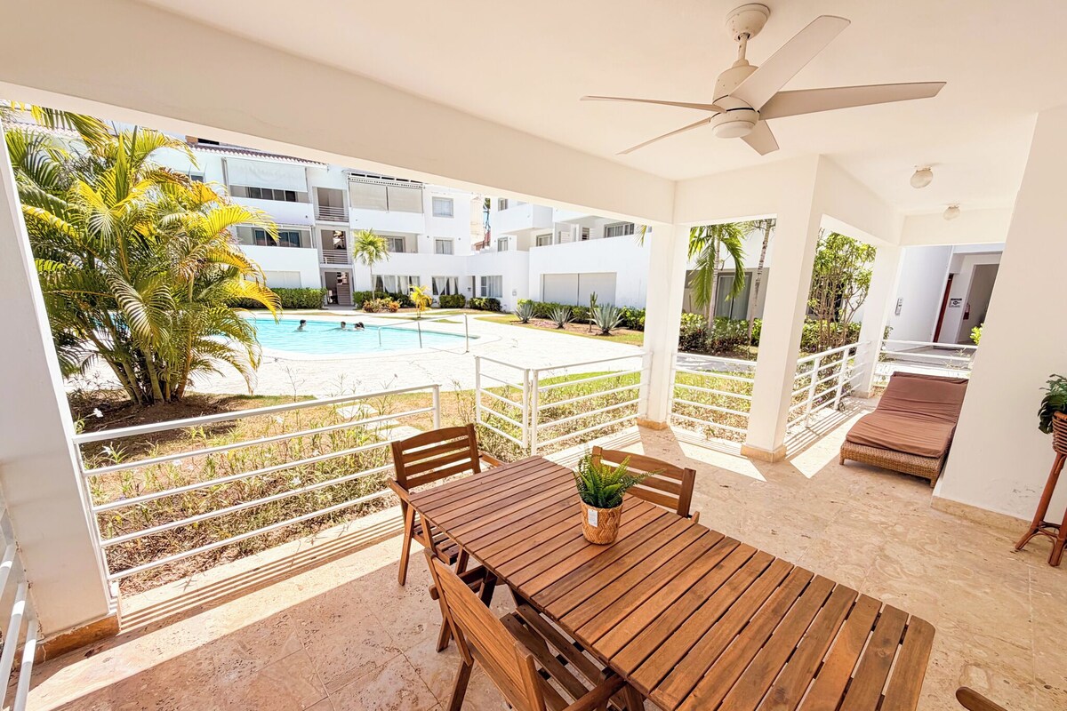 Uma área externa coberta com piscina, móveis de madeira e vista para um edifício residencial.