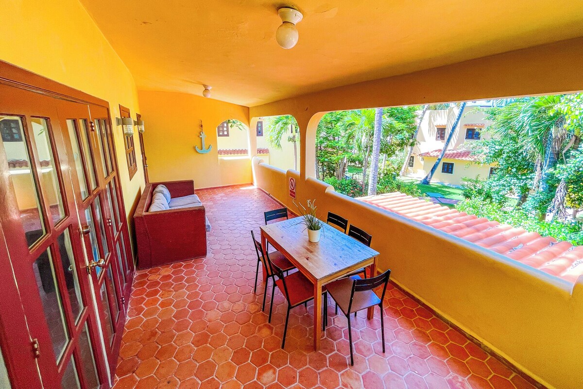 Quarto com piso de azulejos, uma mesa com cadeiras, um sofá e uma janela com vista para um pátio.