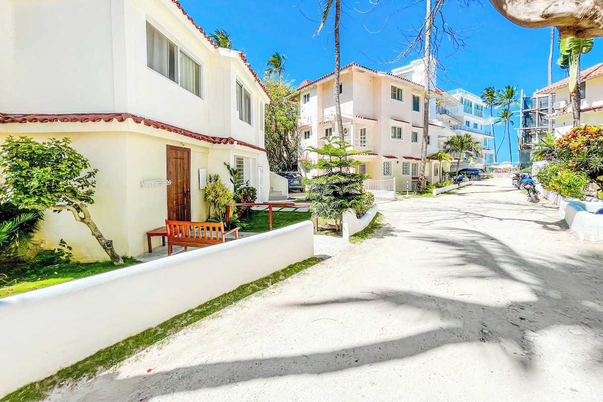 Rua residencial com casas brancas, palmeiras e um banco.