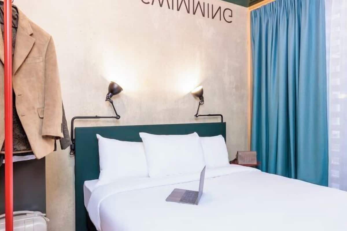 Quarto de hotel com uma cama, um laptop, um cabideiro e uma cortina azul.