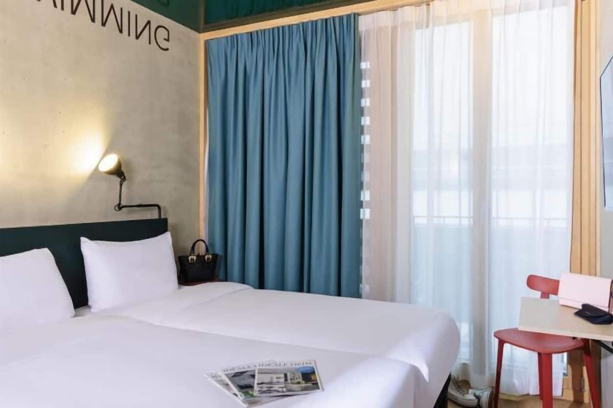Quarto de hotel com uma cama, uma escrivaninha, uma cadeira, uma bolsa e uma revista sobre a cama.