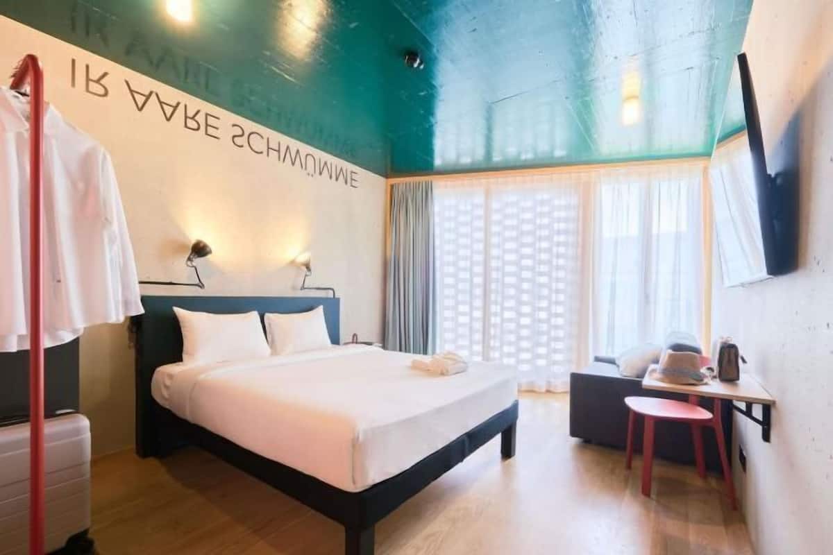 Quarto de hotel com cama, escrivaninha, cadeira, guarda-roupa e janela com cortinas.