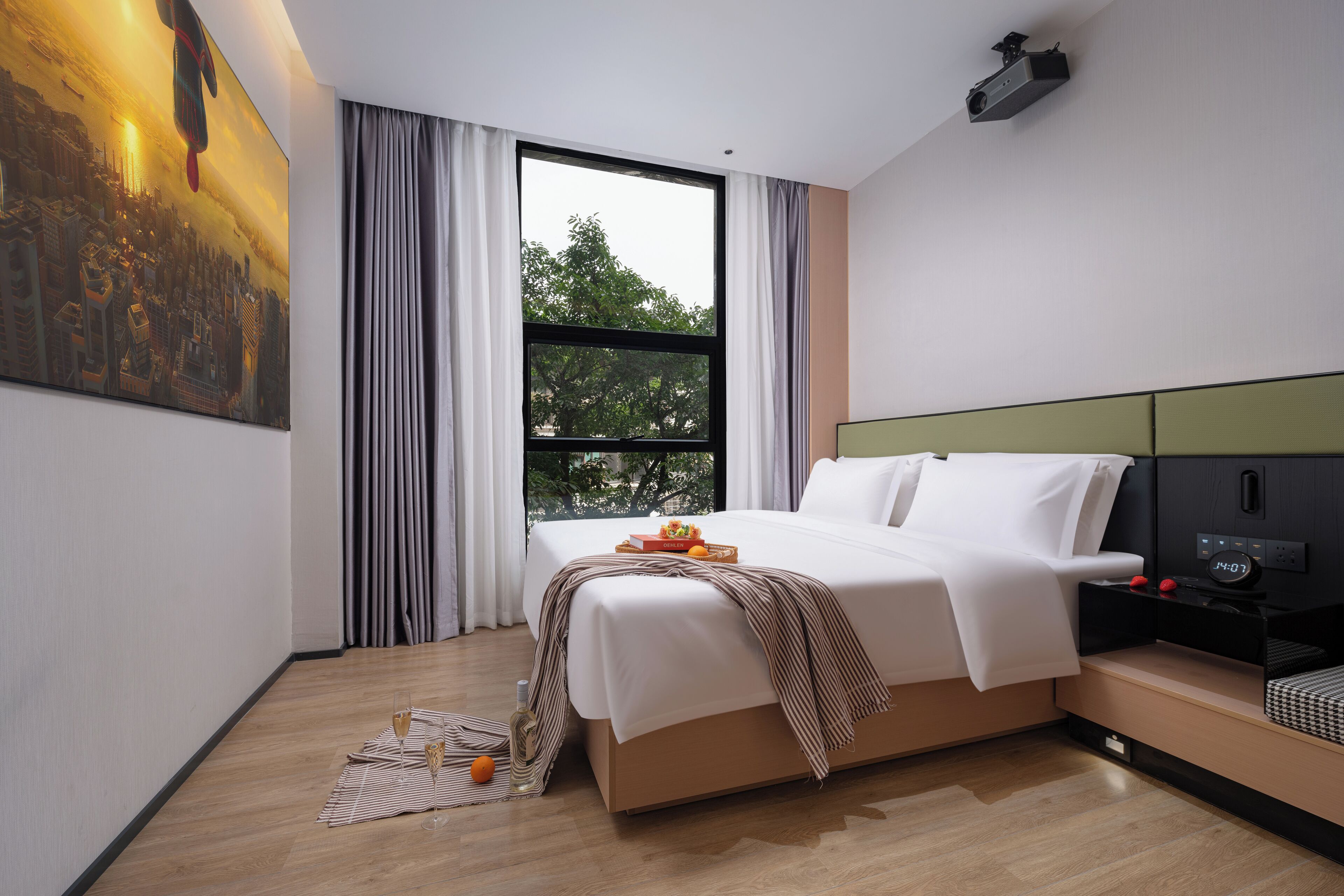 Quarto de hotel moderno com uma cama grande, uma mesa de cabeceira, um projetor e um quadro na parede.