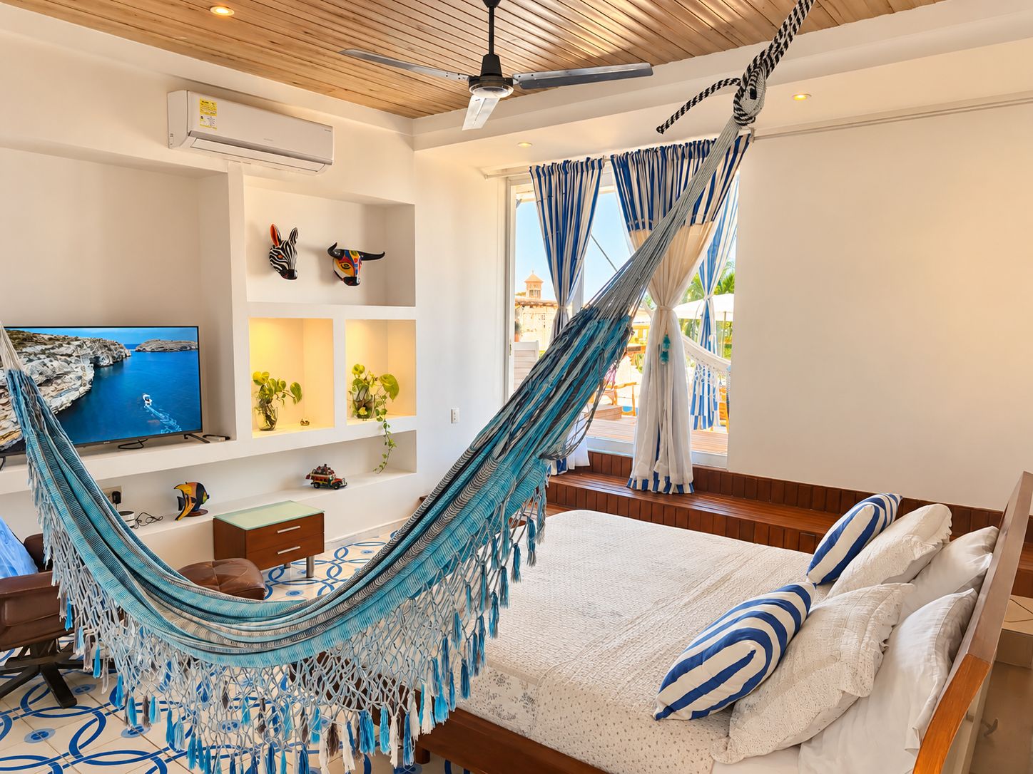 Quarto com rede, cama com almofadas listradas, televisão e objetos decorativos nas prateleiras.