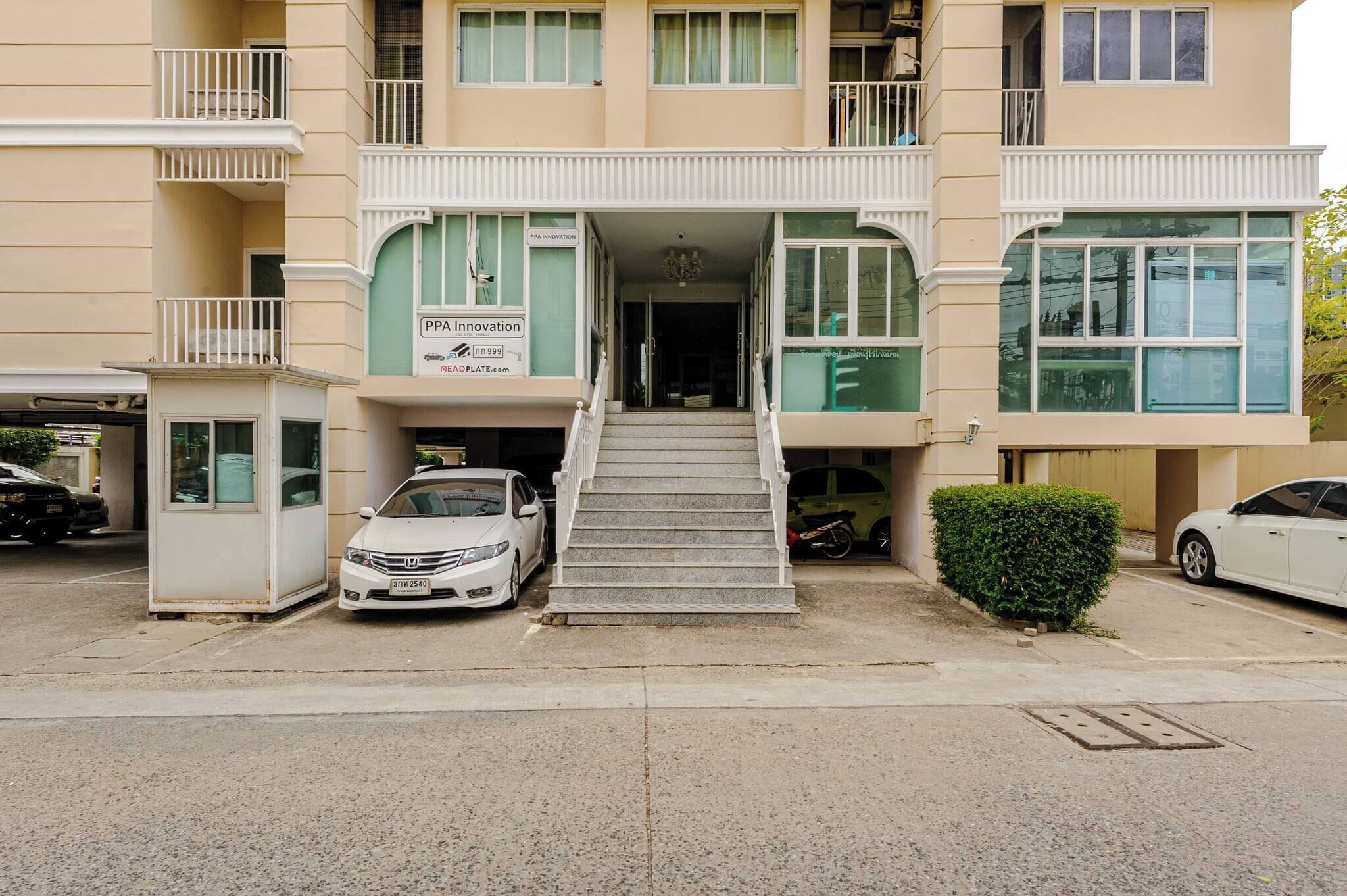 Um edifício de vários andares com entrada central, uma escadaria que leva ao andar térreo e um carro estacionado em frente.