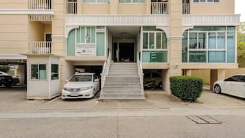 Um edifício de vários andares com entrada central, uma escadaria que leva ao andar térreo e um carro estacionado em frente.