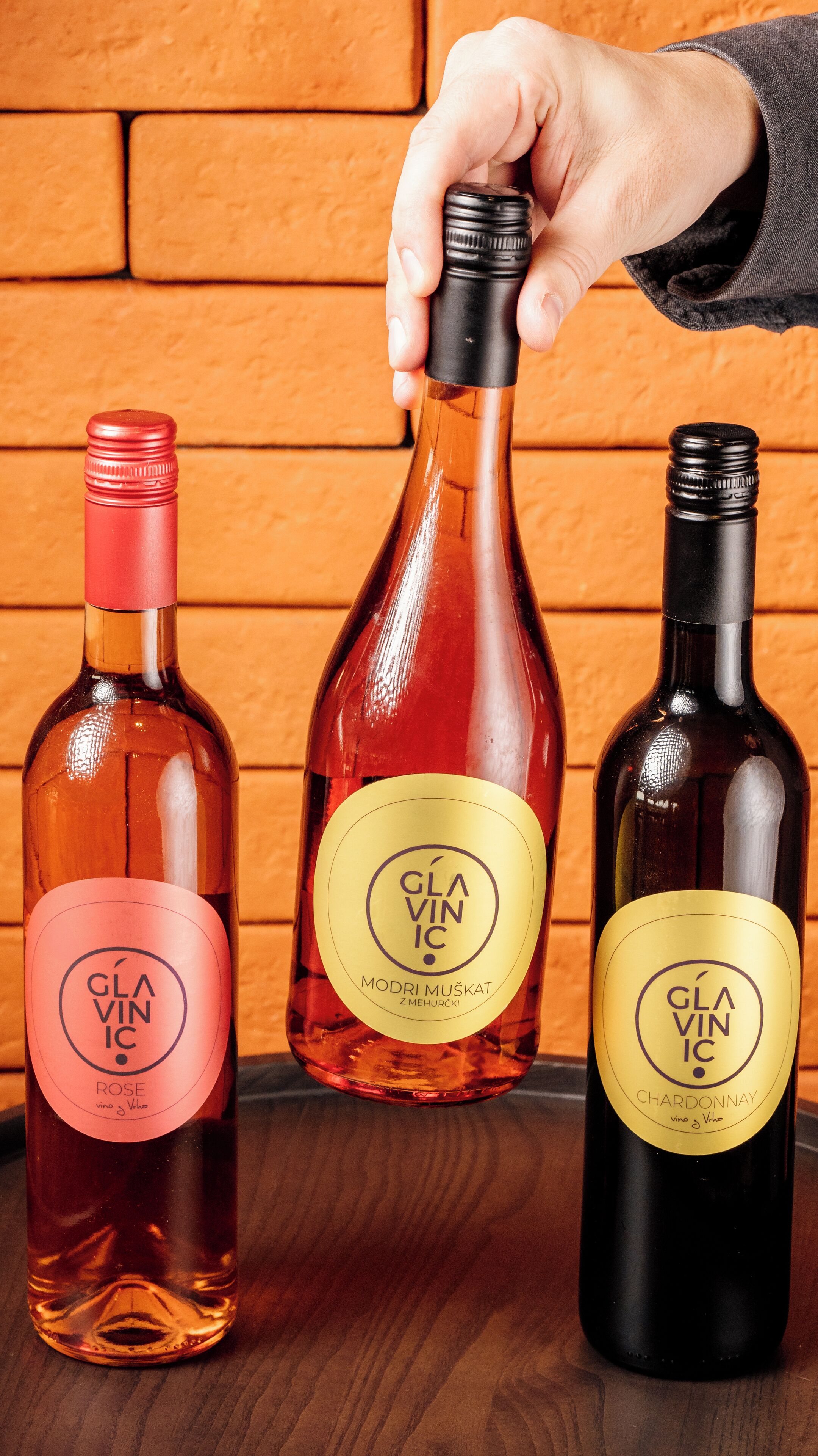 Três garrafas de vinho com etiquetas, uma delas sendo segurada por uma mão, com um fundo de parede de tijolos.