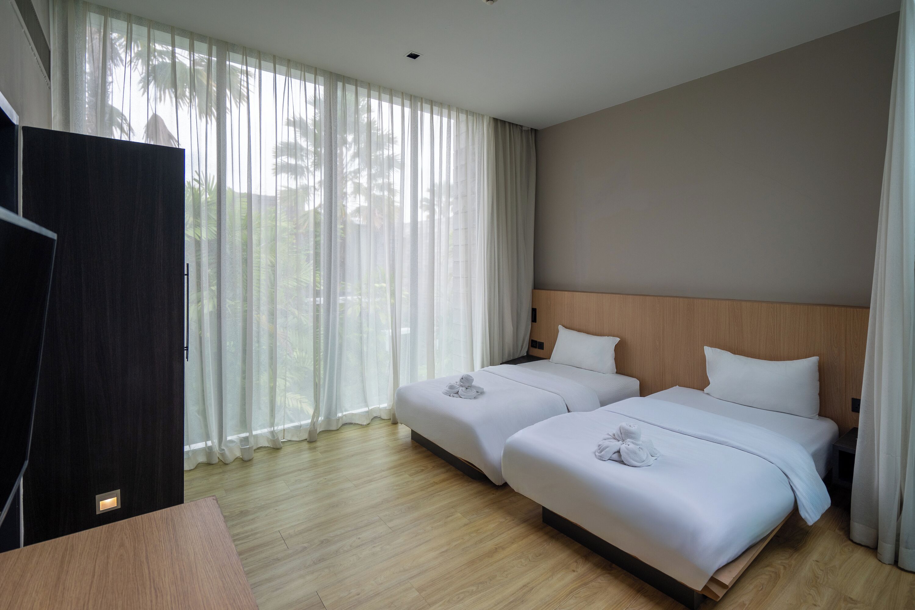 Quarto de hotel com duas camas, cabeceira de madeira, televisão e janelas amplas com cortinas leves.