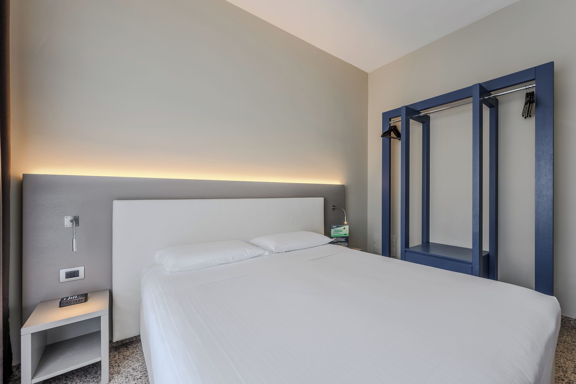 Quarto de hotel com cama, mesa de cabeceira, guarda-roupa e luminária na parede.