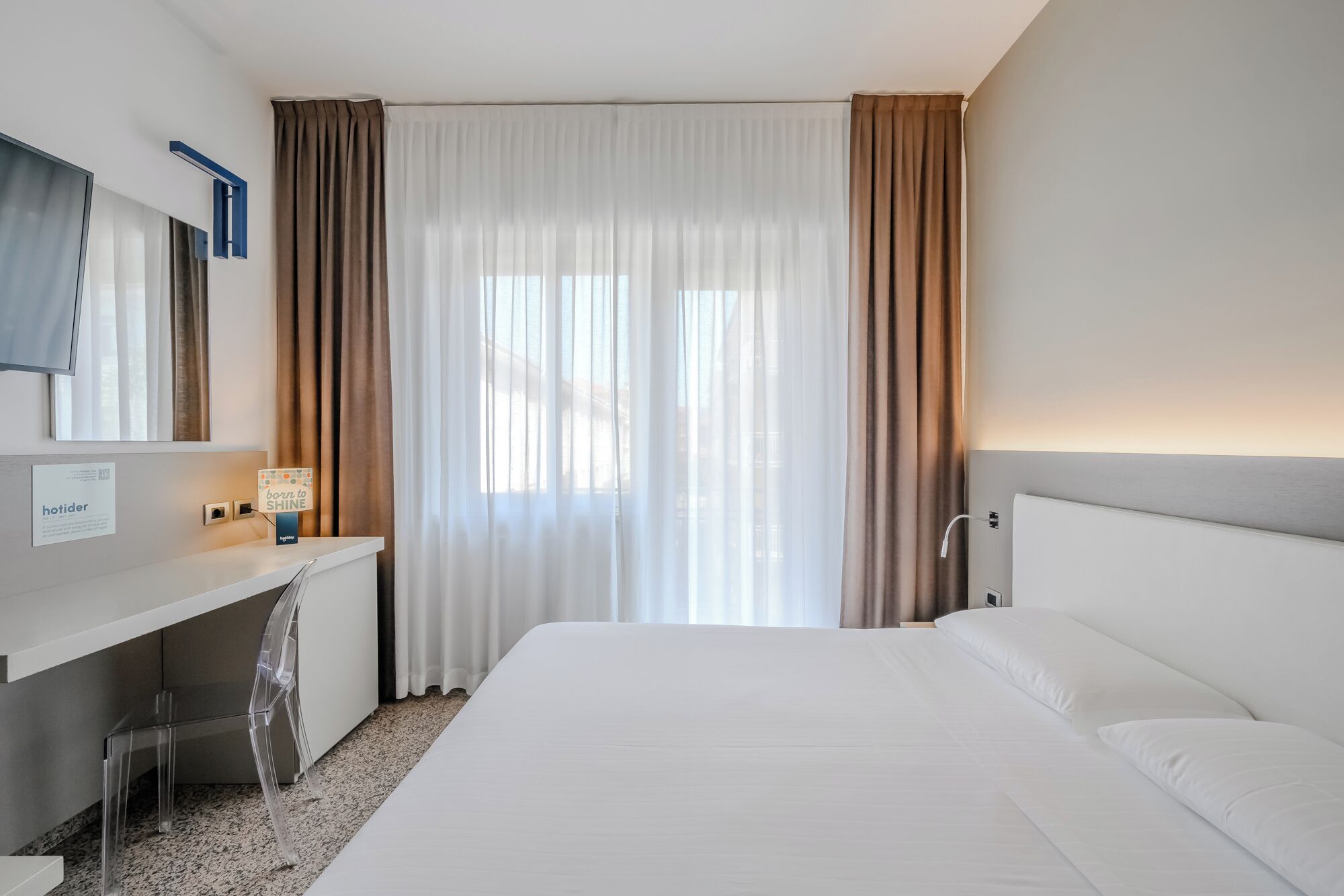 Quarto de hotel com cama, escrivaninha, cadeira, abajur e janela com cortinas.