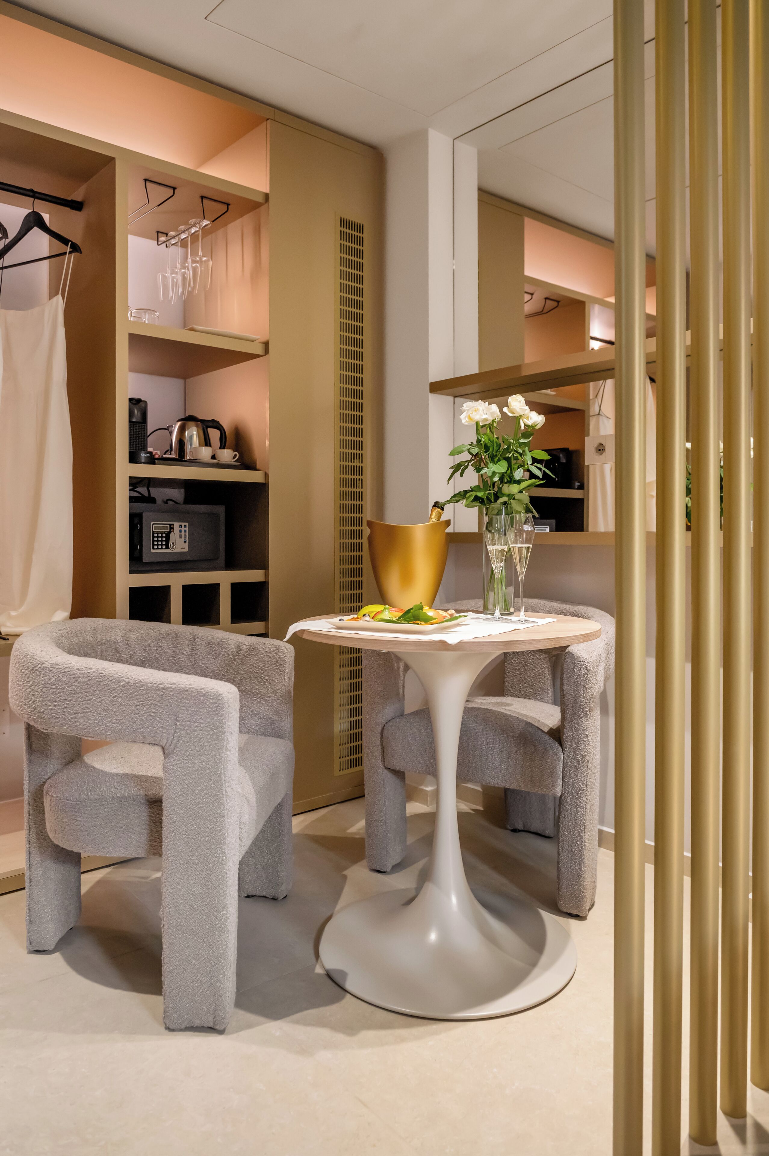 Quarto moderno com uma mesa redonda, duas cadeiras cinzas, um vaso dourado com flores e um micro-ondas ao fundo.