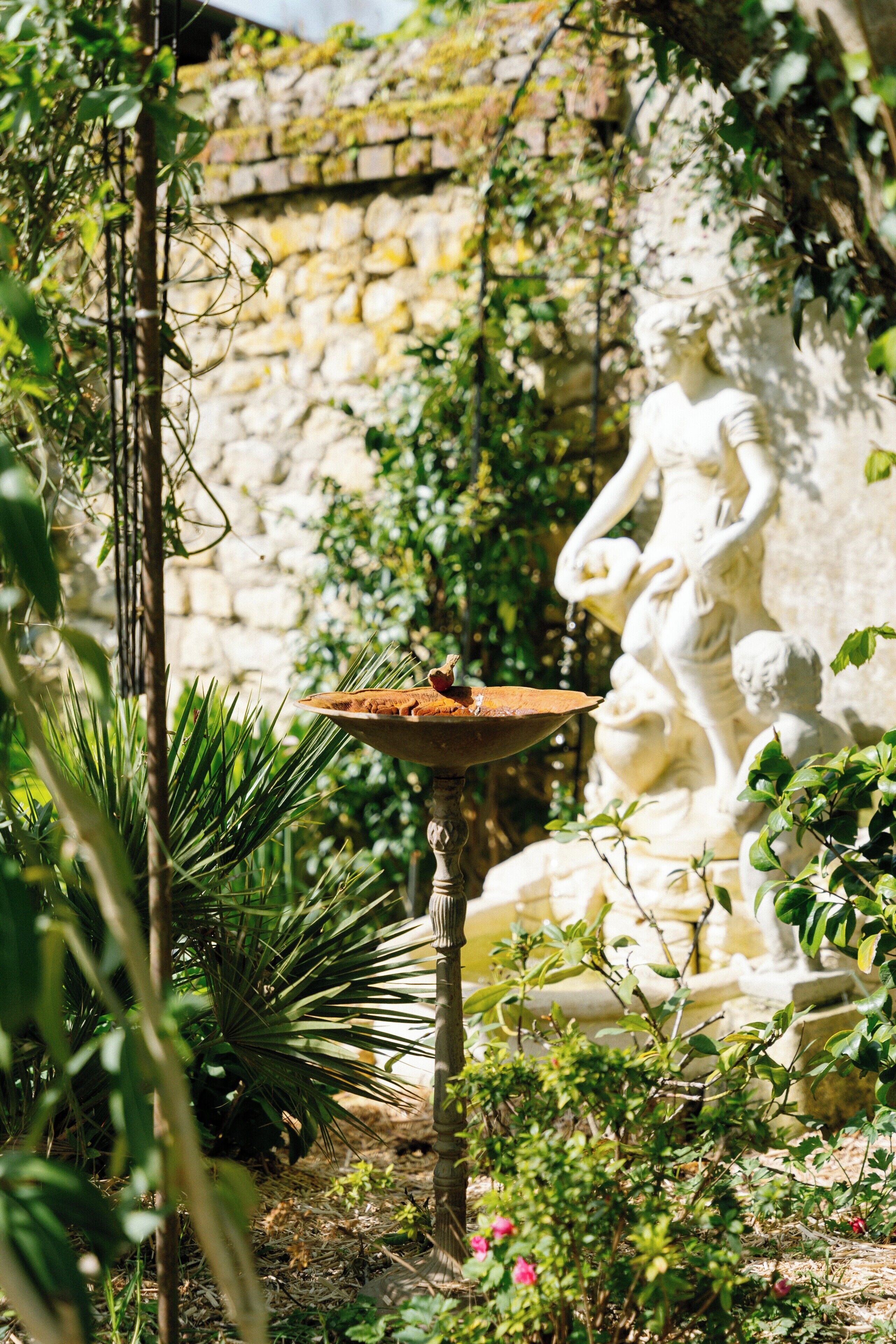 Um jardim com muro de pedra, estátua e bebedouro para pássaros.