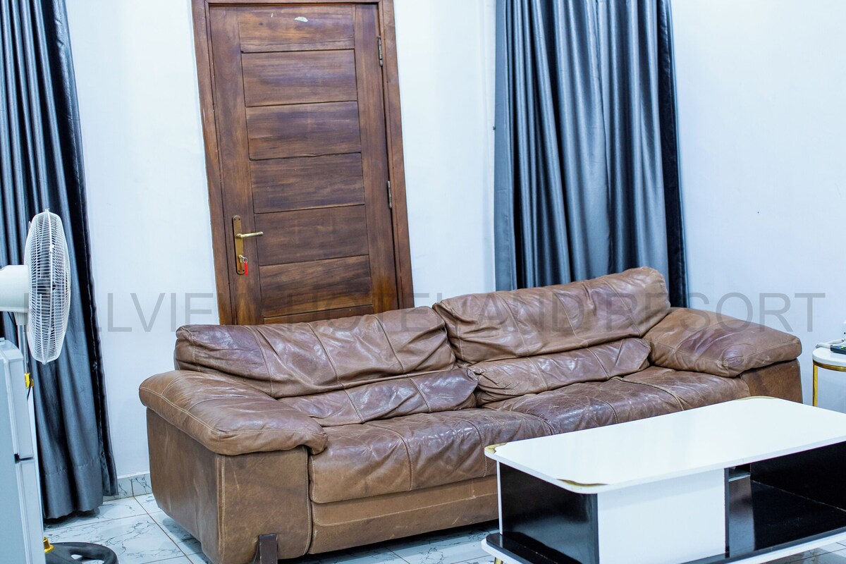 Sala de estar com sofá de couro marrom, mesa de centro branca, porta de madeira e ventilador de teto.