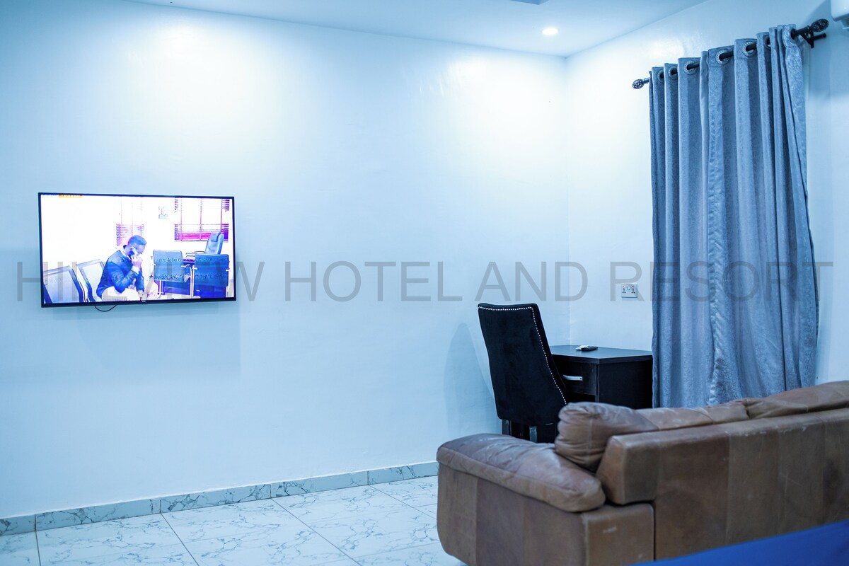 Quarto de hotel com televisão de tela plana na parede, um sofá, uma mesinha e uma cortina.