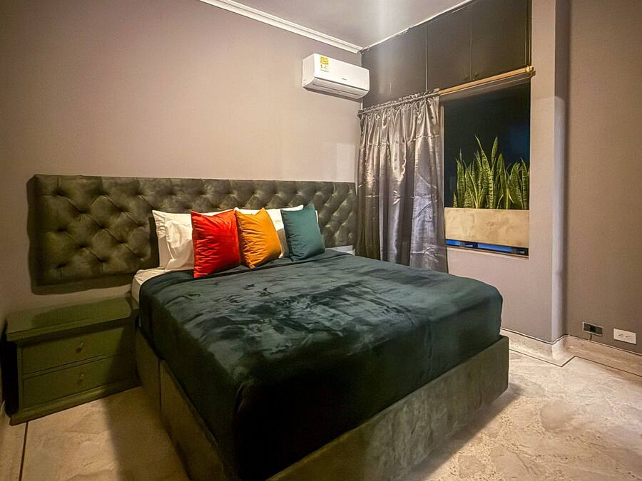 Quarto com cabeceira estofada, cama com colcha verde escura, almofadas coloridas, mesa de cabeceira e janela com cortinas.