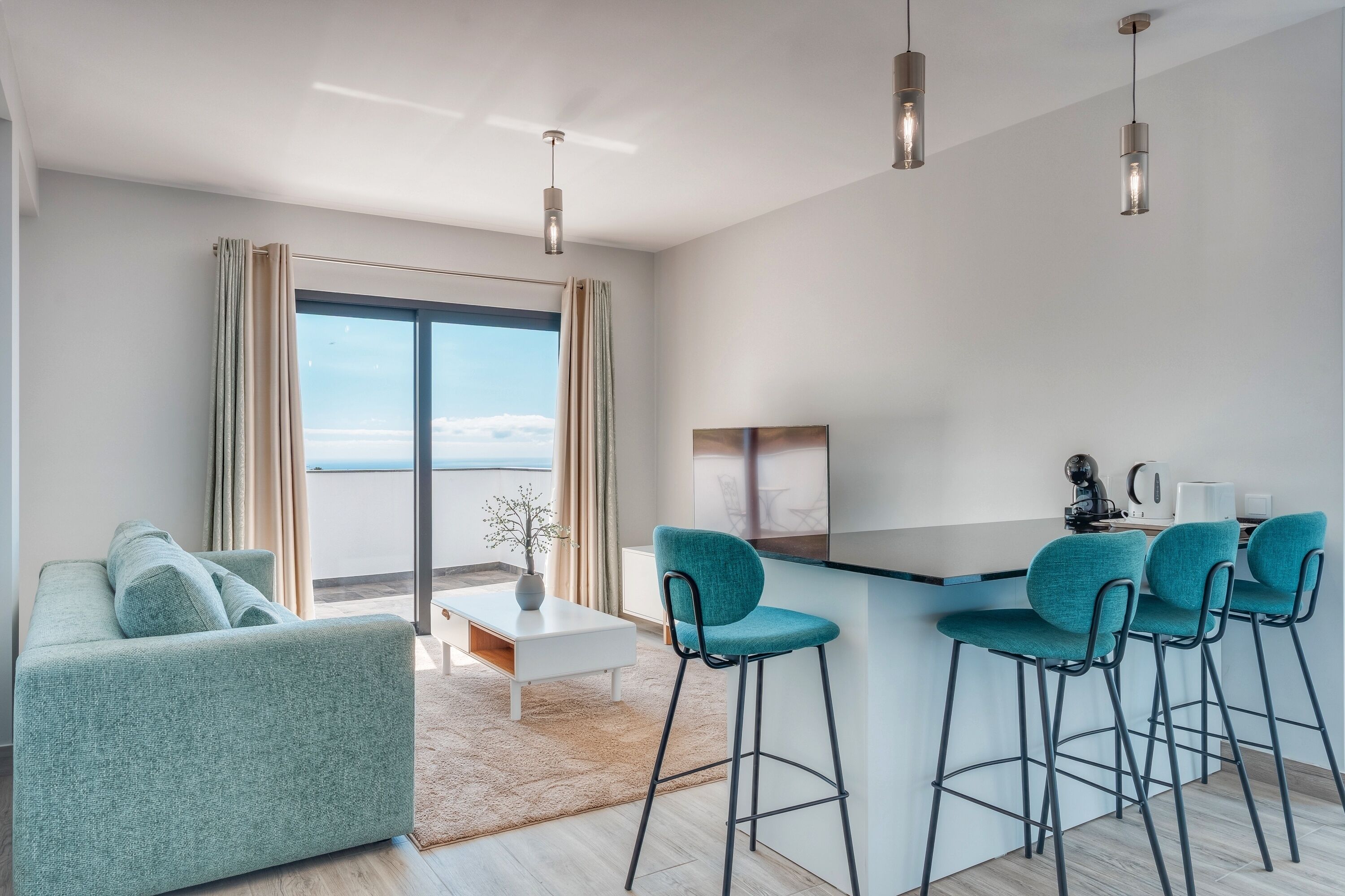 Sala de estar moderna com sofá e mesa de centro, área de cozinha com banquetas e uma janela grande com cortinas.