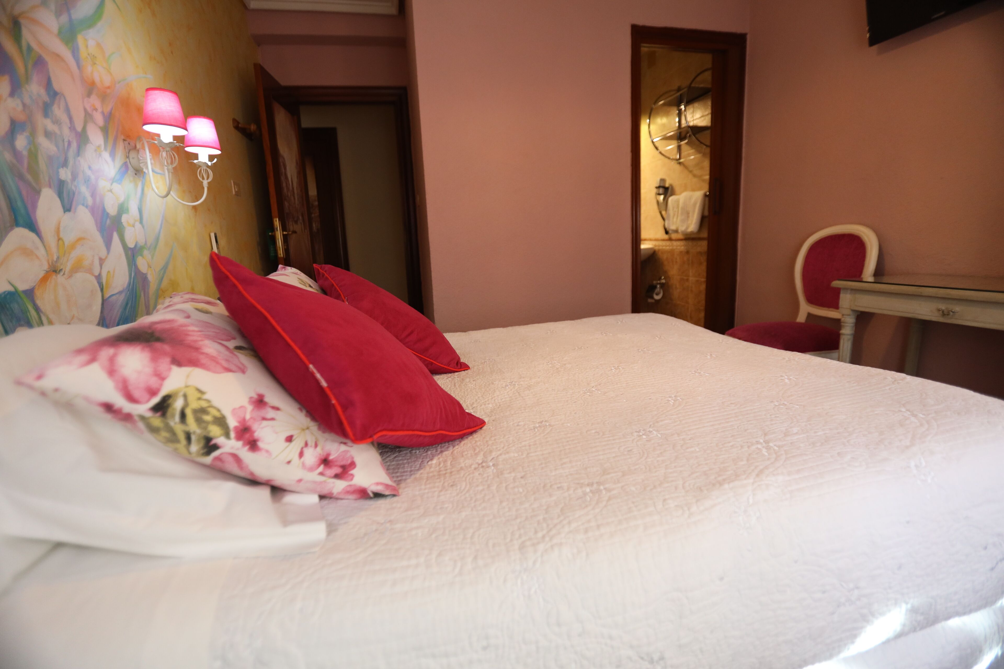 Cama bem arrumada com almofadas vermelhas e uma almofada com padrão floral, uma mesinha de cabeceira e uma luminária fixada na parede.