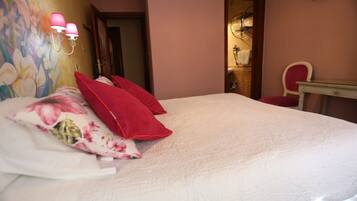 Cama bem arrumada com almofadas vermelhas e uma almofada com padrão floral, uma mesinha de cabeceira e uma luminária fixada na parede.