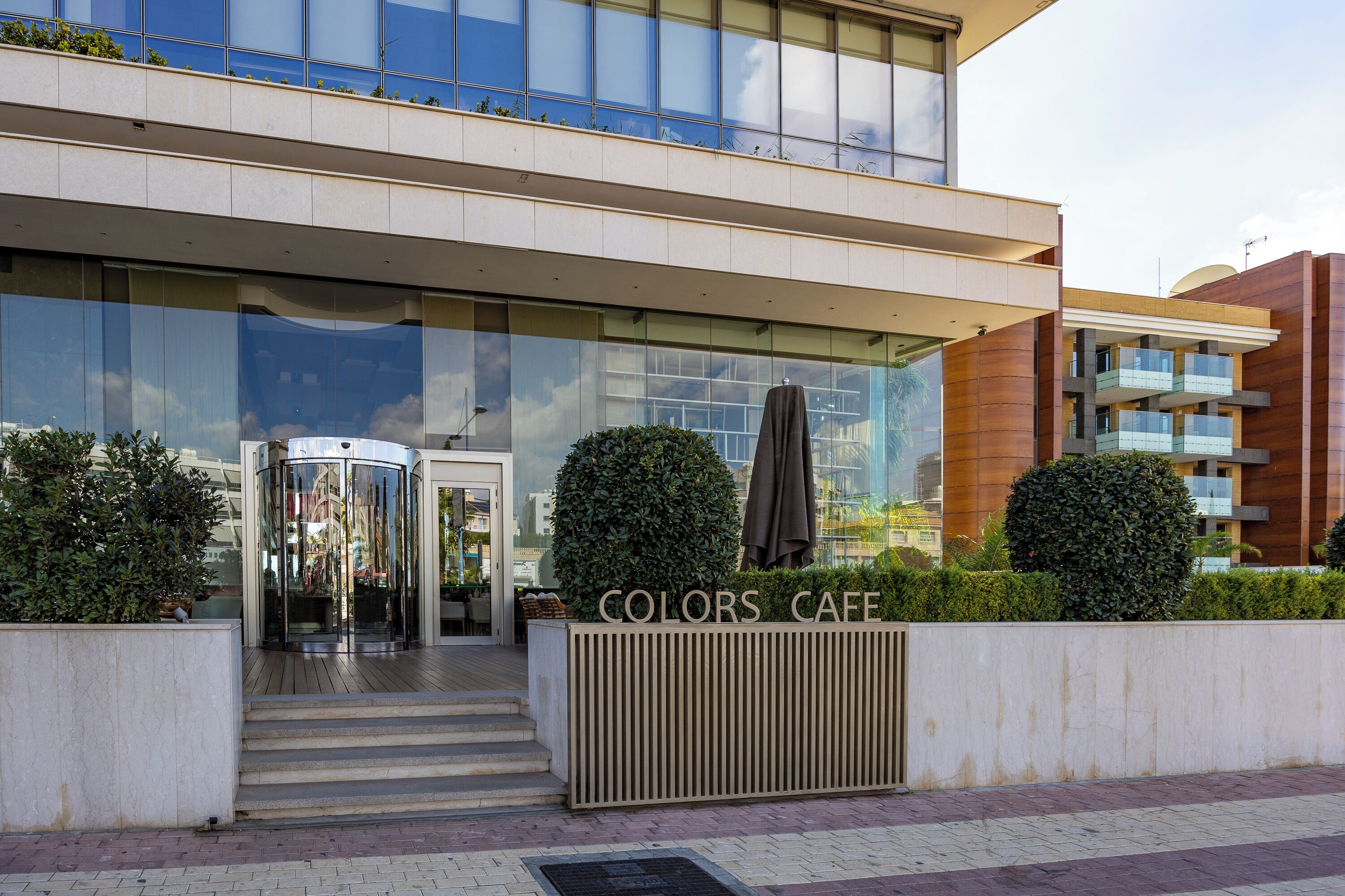 Um edifício moderno com entrada de vidro, uma placa com a inscrição "COLORS CAFE" e uma pequena área de estar ao ar livre com degraus que levam à entrada.