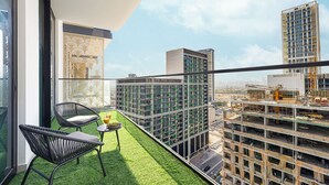 Uma varanda com grama sintética, duas cadeiras de vime, uma mesa pequena com uma cesta de frutas e vista para um horizonte urbano com edifícios altos.