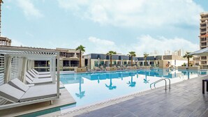 Piscina com espreguiçadeiras e guarda-sóis, rodeada por palmeiras e edifícios modernos.