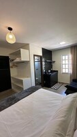 Quarto de hotel com cama, escrivaninha, armário, chuveiro e janela.