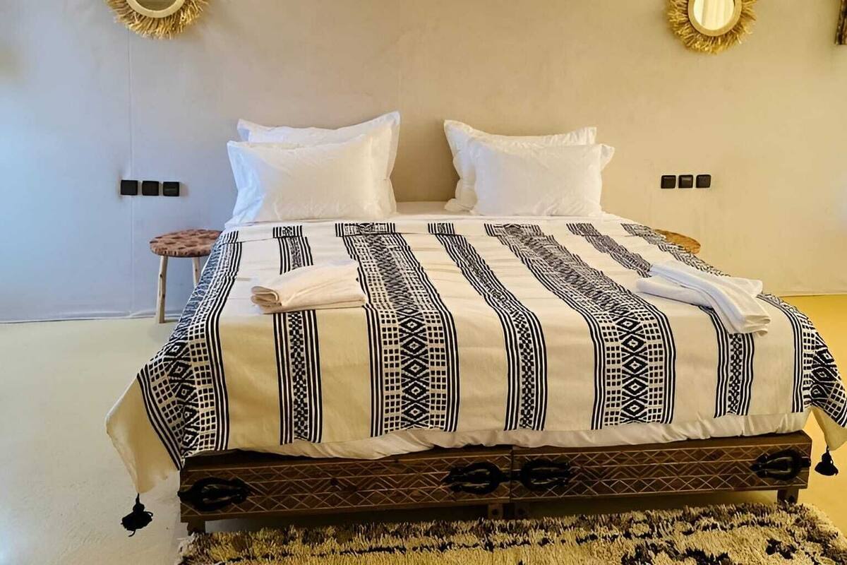 Uma cama com colcha estampada, duas almofadas e cabeceira. Há duas arandelas com espelhos redondos.