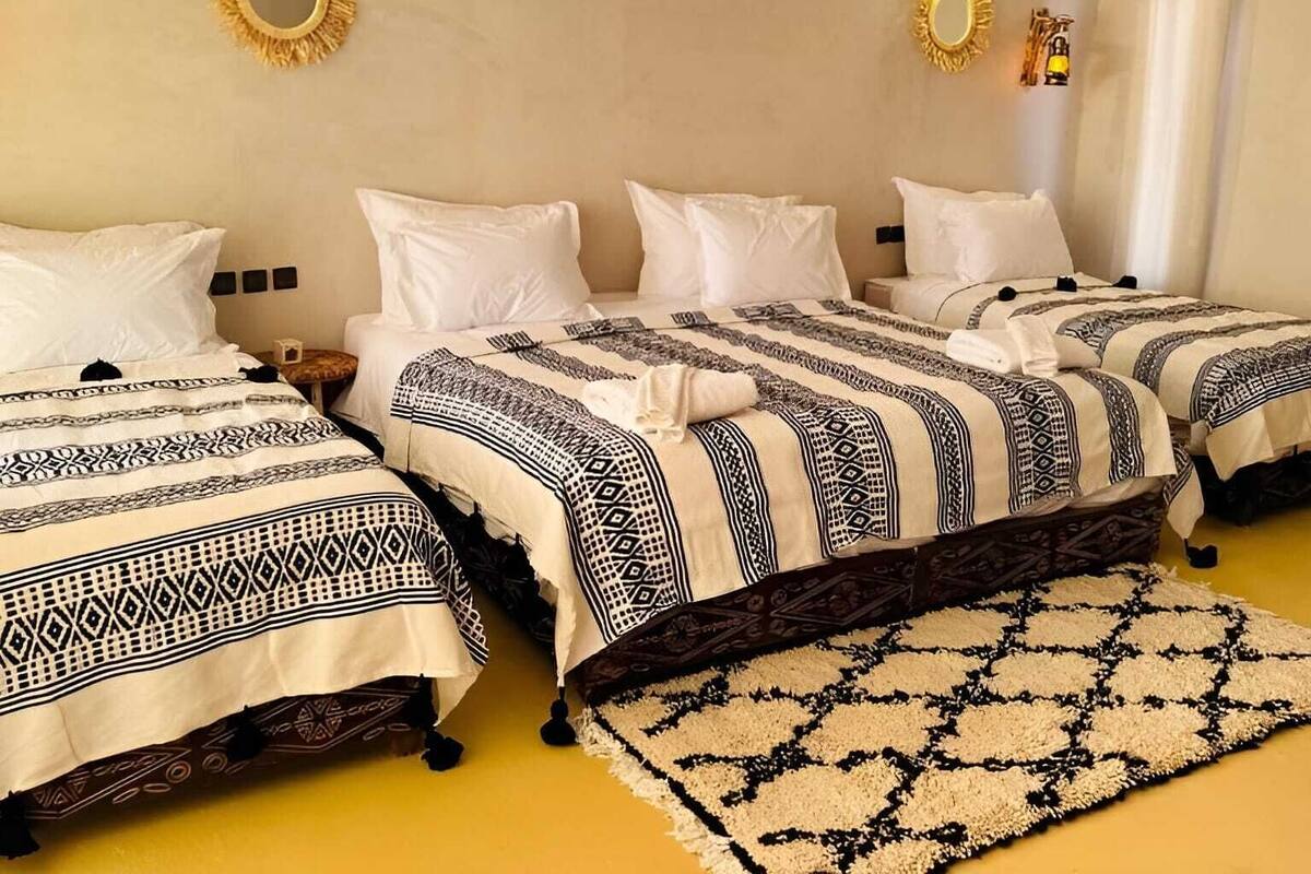 Quarto de hotel com três camas, cada uma com roupa de cama estampada, um espelho redondo na parede e uma pequena mesa com abajur.