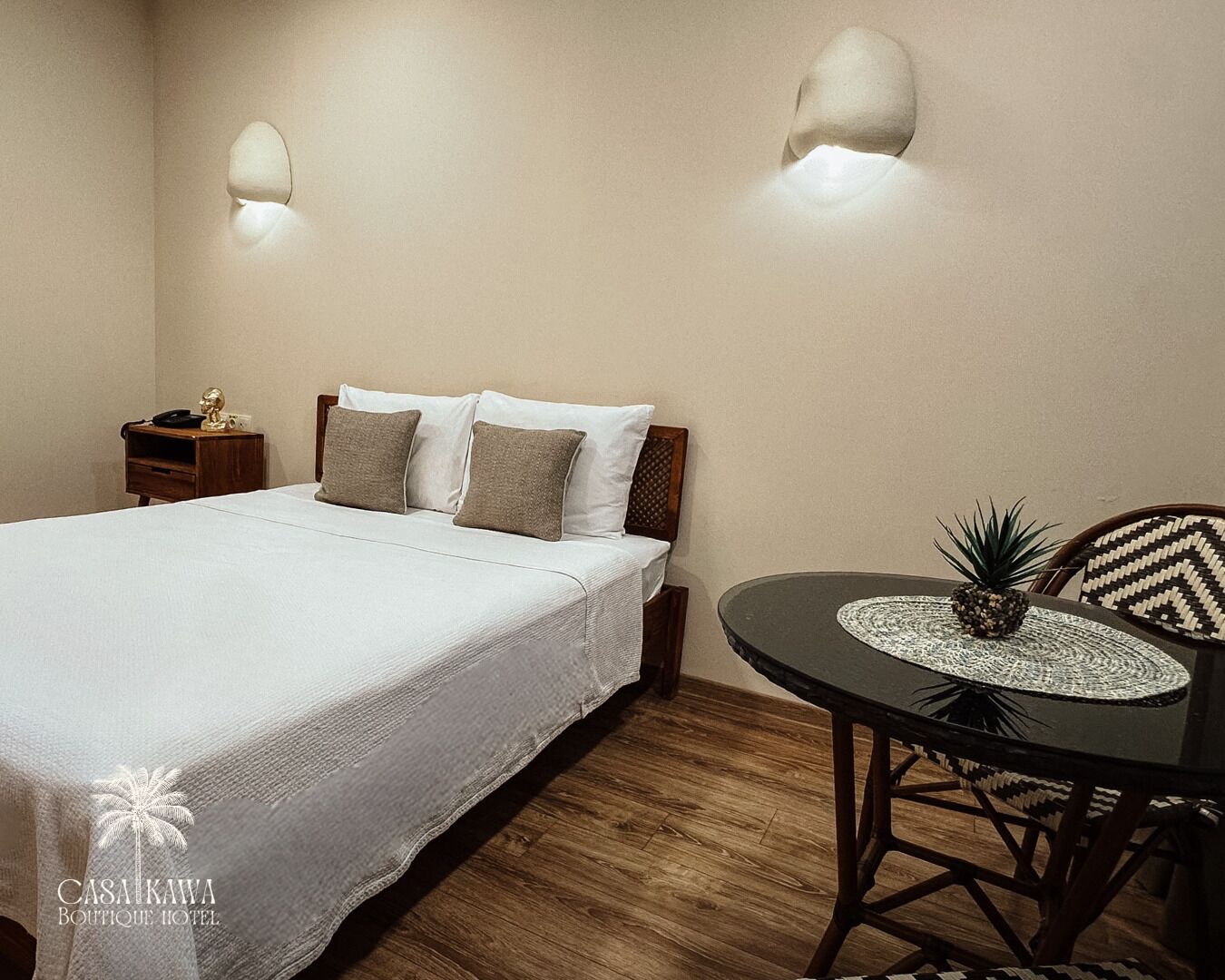 Quarto com cama, mesa de cabeceira, luminária e uma pequena mesa redonda com decoração de abacaxi.