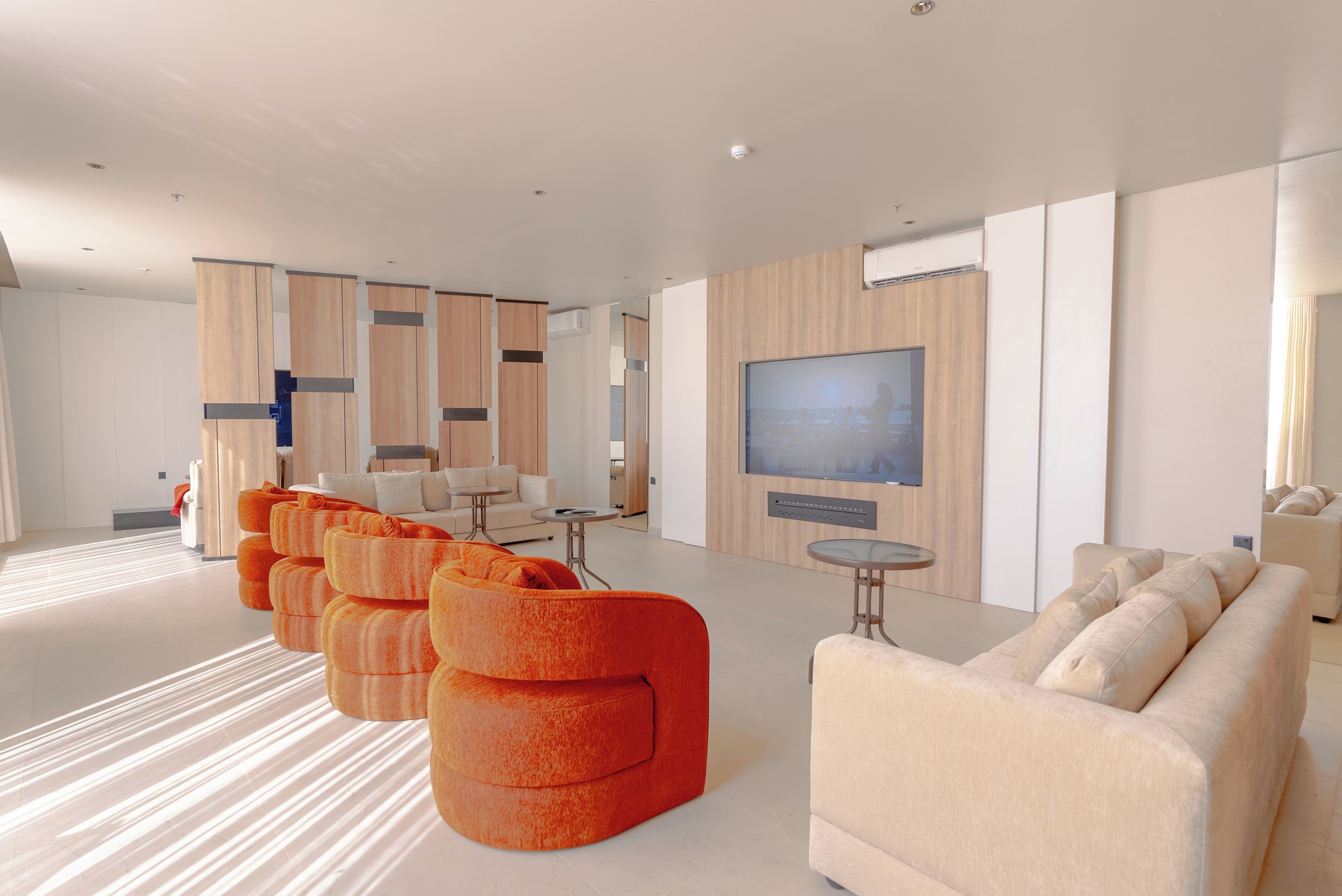 Uma sala de estar moderna com uma televisão de tela plana grande, diversas opções de assento incluindo sofás laranja e bege, e um design minimalista com detalhes em madeira.
