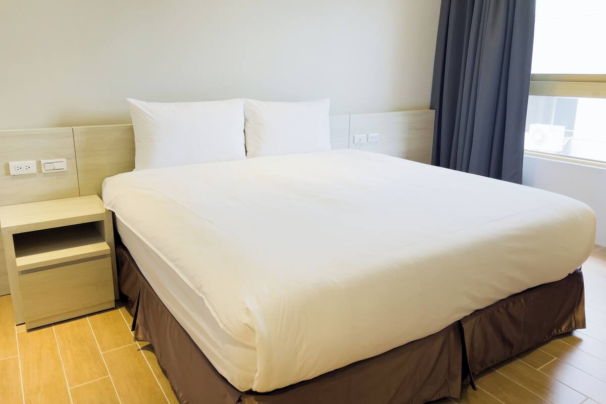 Quarto de hotel com cama grande, mesa de cabeceira, cômoda e janela com cortinas.