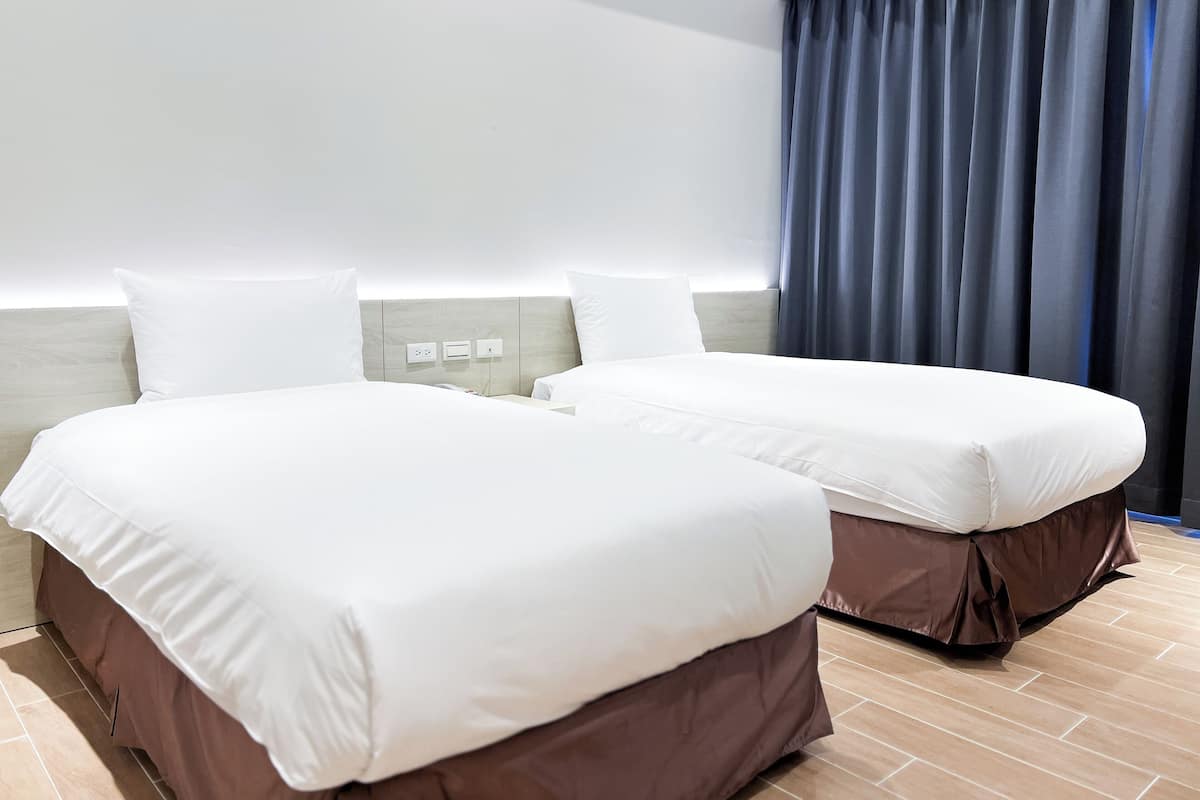 Quarto de hotel com duas camas, cabeceira de madeira, mesa de cabeceira e uma janela grande com cortinas escuras.