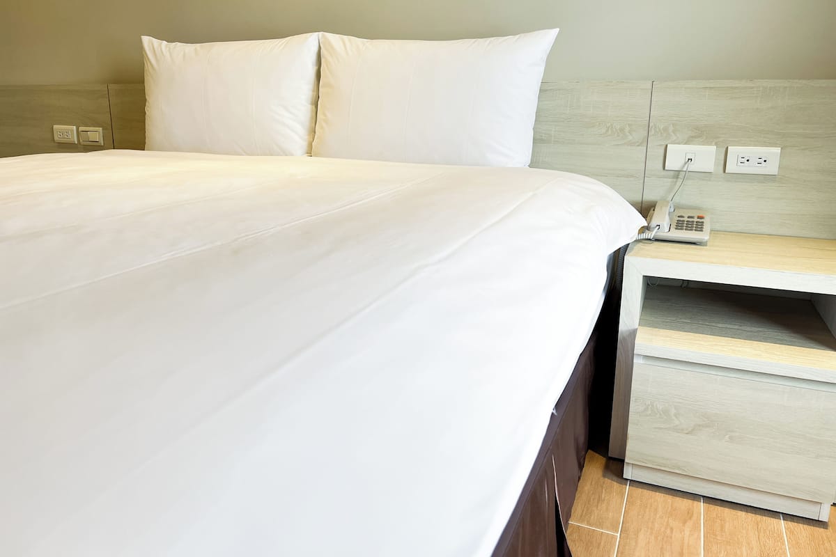 Cama bem arrumada com lençóis brancos, mesa de cabeceira com telefone e tomada na parede.
