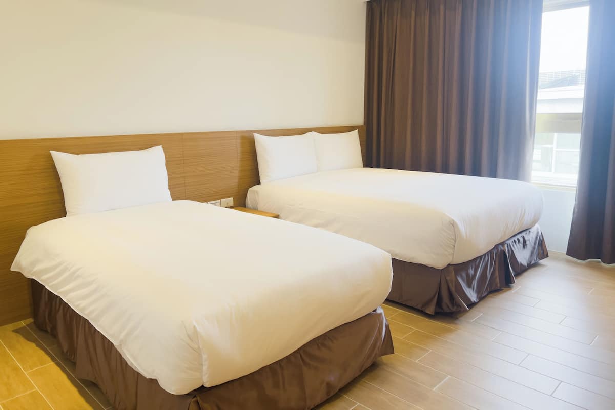Quarto de hotel com duas camas, cabeceiras de madeira, roupa de cama branca e uma janela grande com cortinas.
