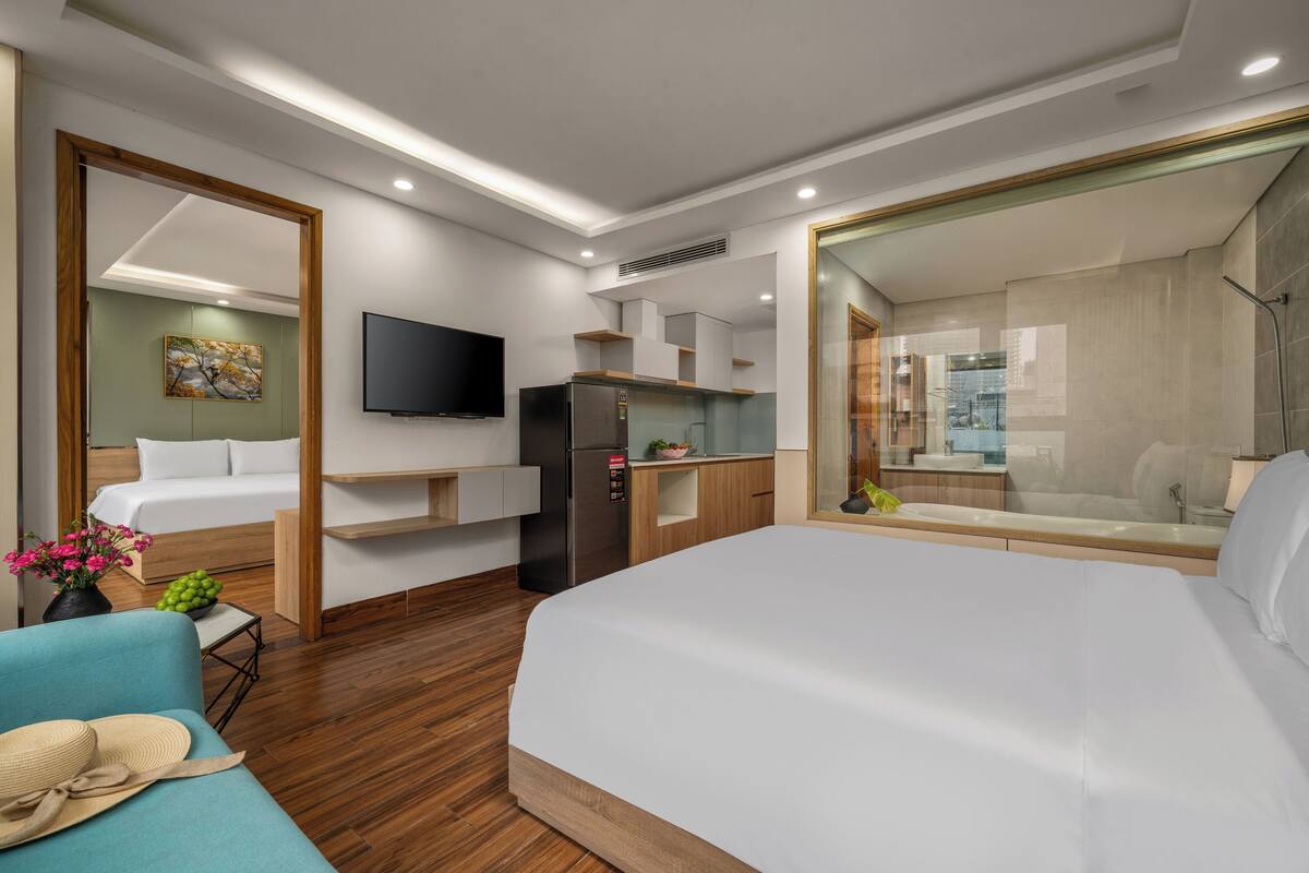 Quarto de hotel moderno com uma cama grande, televisão de tela plana, cozinha compacta e banheiro com paredes de vidro.