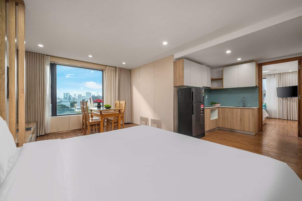 Quarto de hotel com uma cama grande, uma mesa de jantar com cadeiras, uma área de cozinha com frigorífico e uma televisão num canto.