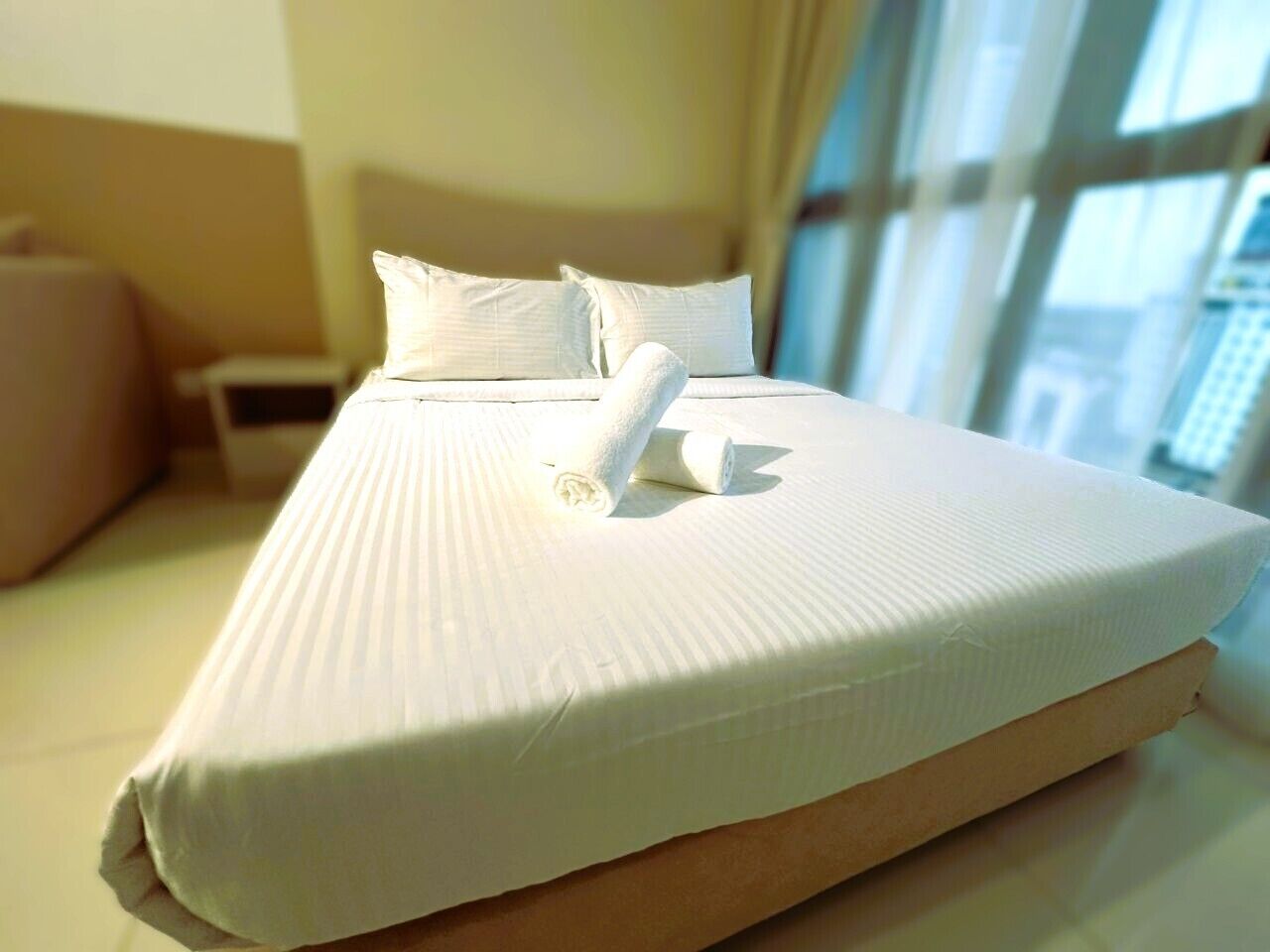 Uma cama bem arrumada com roupa de cama branca e uma toalha enrolada, uma mesa de cabeceira e uma janela com vista para edifícios.